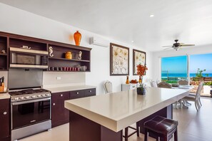 Uma cozinha moderna com ilha central, forno embutido e micro-ondas. Há uma área de jantar com vista para o oceano.