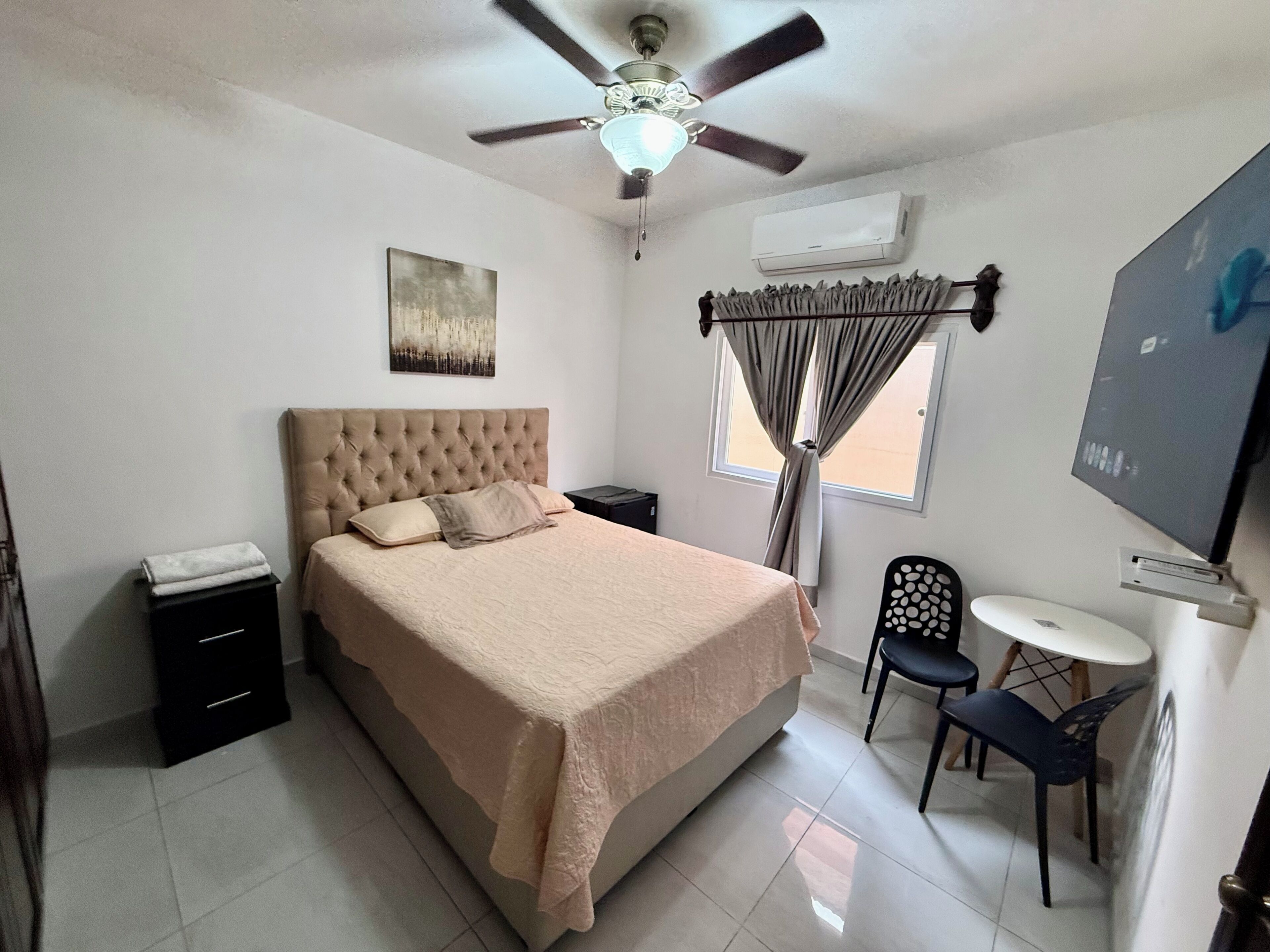 Quarto com cama, ventoinha no teto, ar condicionado, uma mesinha com cadeira e televisão.