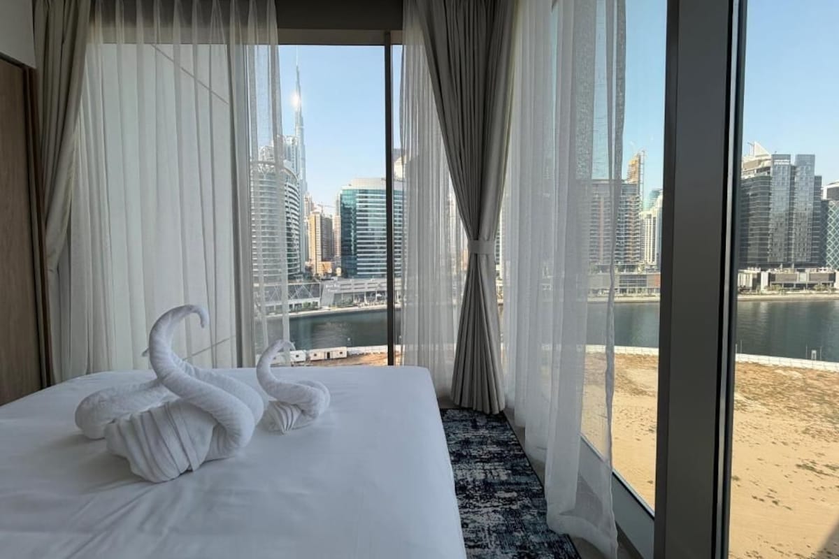 Quarto de hotel com cama, cortinas leves e vista para o horizonte urbano e para a praia.