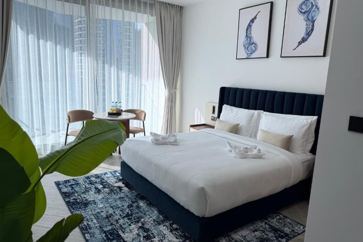 Um quarto com cama, mesa de cabeceira, cadeira, janela com cortinas e duas obras de arte emolduradas na parede.