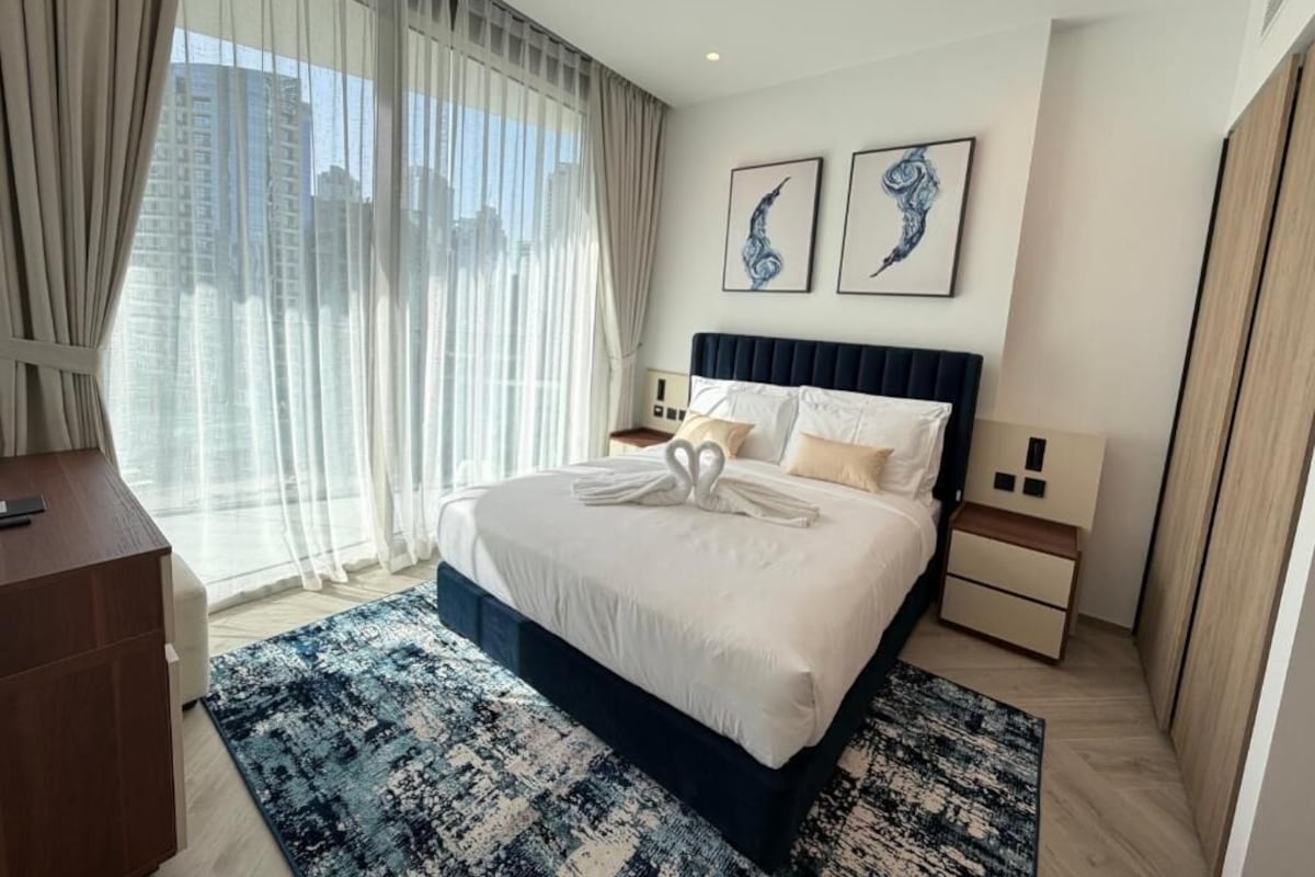 Quarto de hotel com uma cama grande, escrivaninha, mesa de cabeceira e vista para a cidade através de cortinas leves.