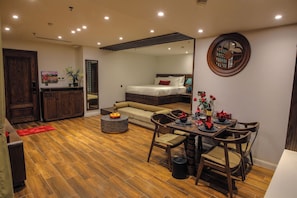 Quarto de hotel moderno com piso de madeira, um sofá, uma mesa de jantar com cadeiras, uma cama, um espelho e uma pequena mesa decorativa com flores.
