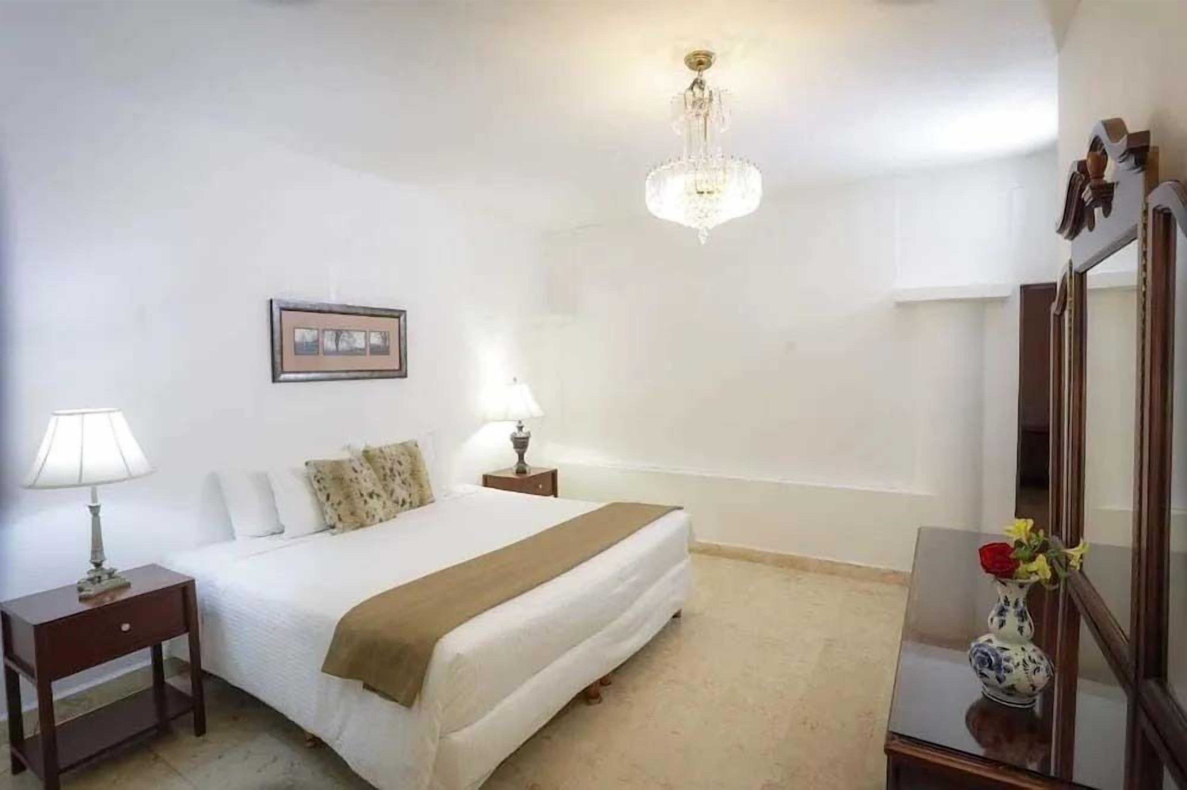 Quarto de hotel com cama, luminárias de cabeceira, um candelabro, um espelho e um vaso com flores.