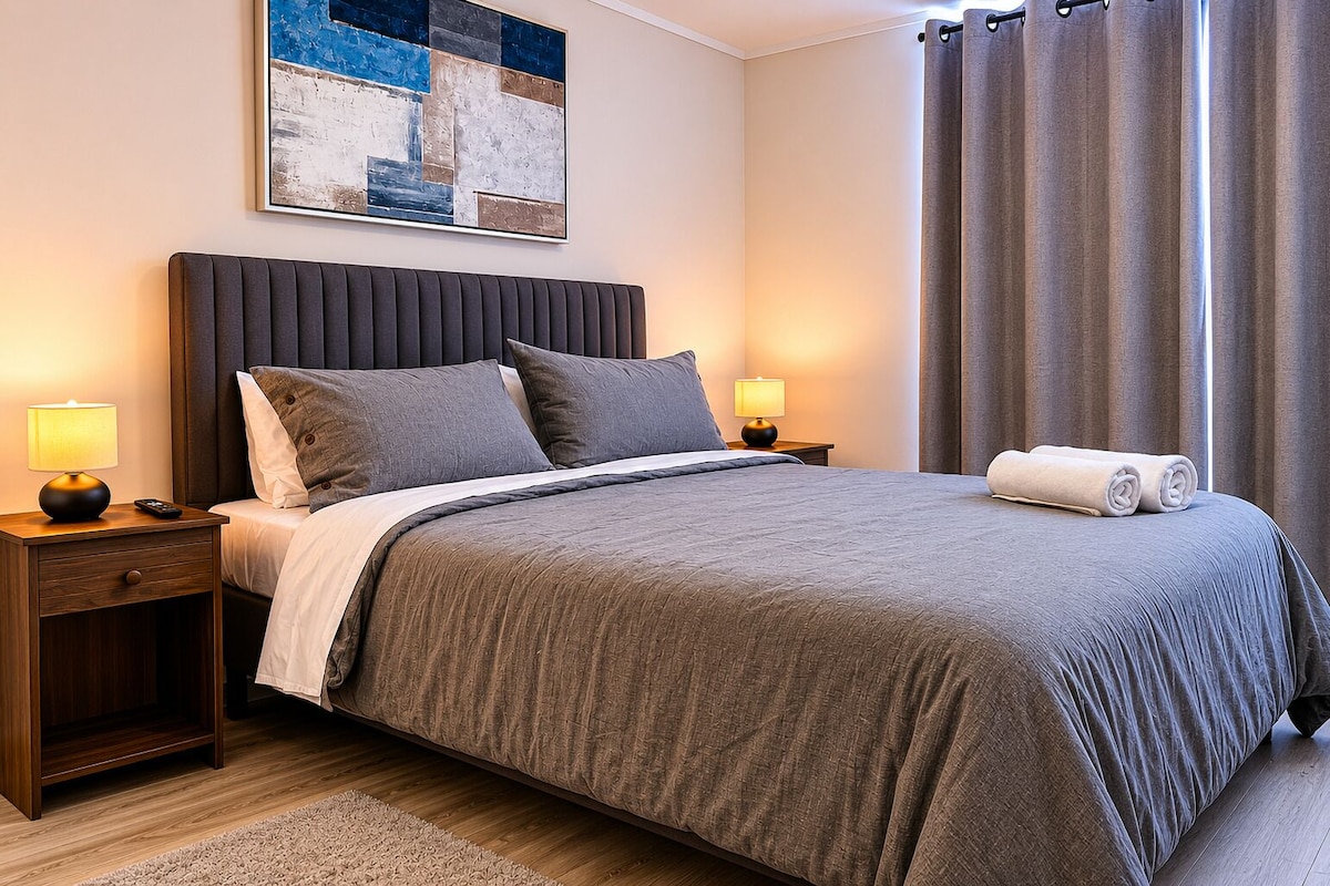 Uma cama bem arrumada com um edredom cinza, duas almofadas, uma mesinha de cabeceira de madeira com um abajur e um controle remoto.