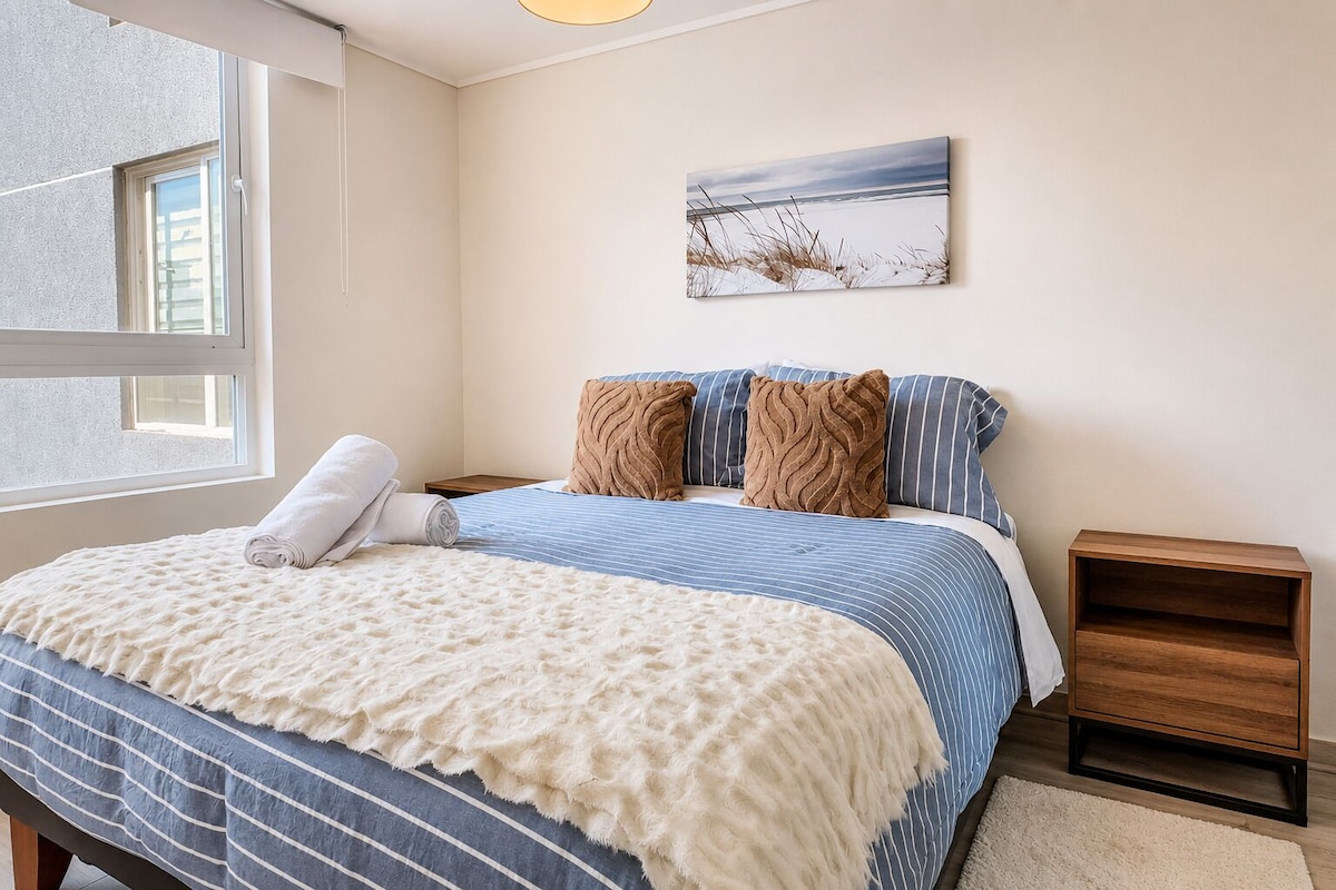 Quarto com cama, almofadas, mesa de cabeceira, janela e um quadro na parede.