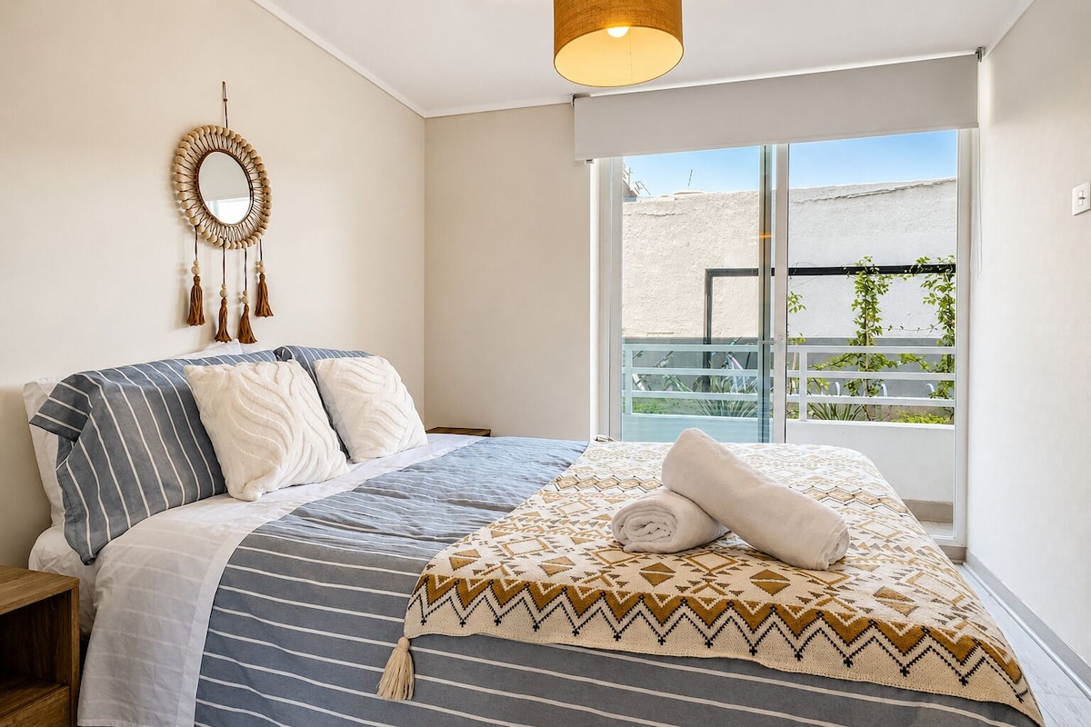 Quarto com cama, espelho fixado na parede e vista para uma varanda com plantas.