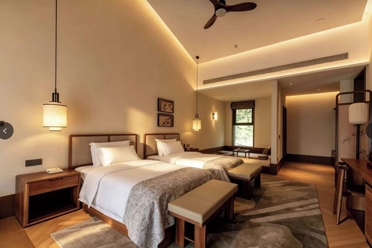 Quarto de hotel com duas camas, uma escrivaninha, uma cadeira, um ventilador de teto e uma janela grande.