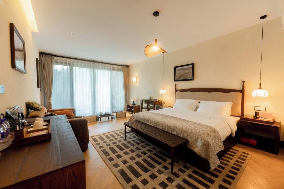 Quarto de hotel com cama, mesas de cabeceira, escrivaninha, cadeira, banco e uma grande janela com cortinas.