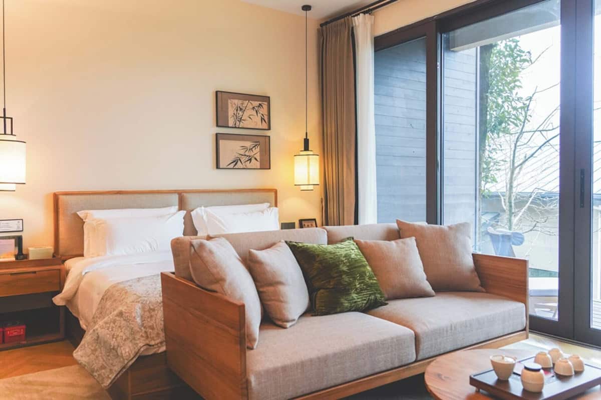 Quarto de hotel com cama, sofá, mesa de cabeceira, abajur e janela com vista para o verde.