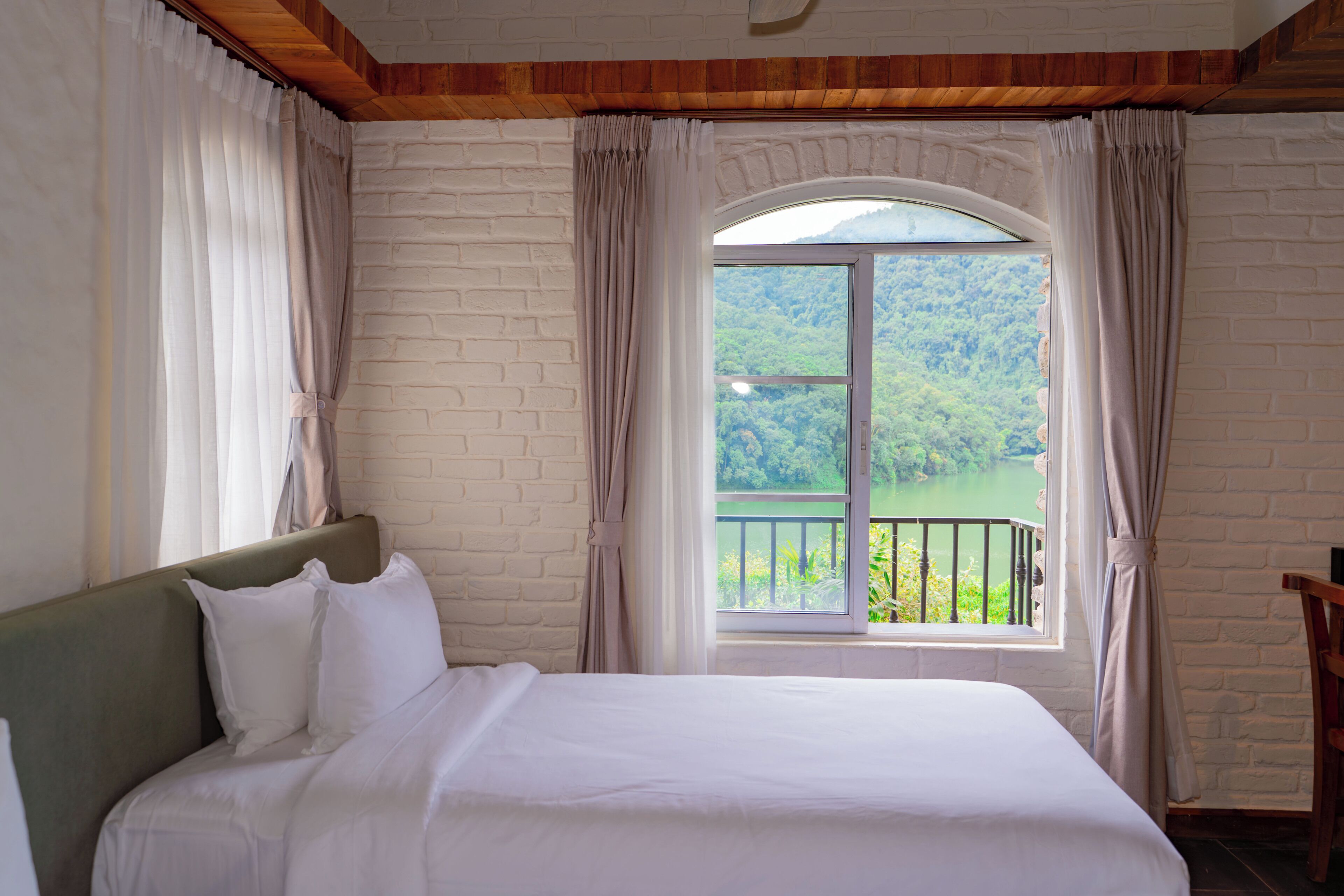 Quarto com cama, janela com cortinas e vista para uma área arborizada.