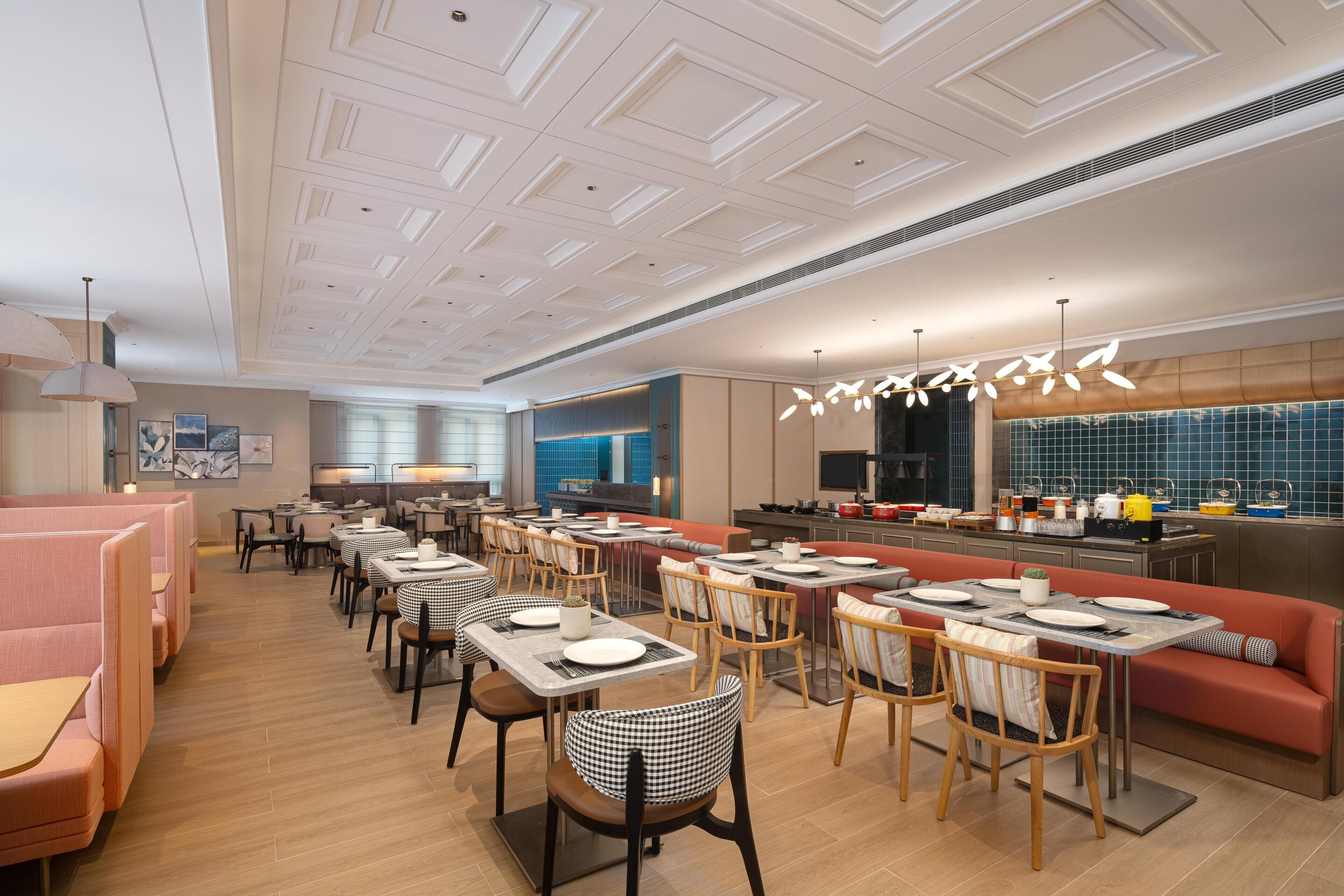 Um restaurante moderno com diversas opções de assentos, incluindo boxes e mesas, uma área de cozinha visível ao fundo e um teto com padrão geométrico.
