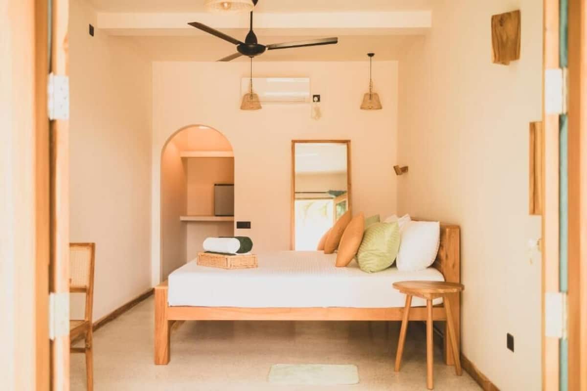 Quarto com cama, ventilador de teto, um banquinho pequeno, um espelho e uma porta em arco.