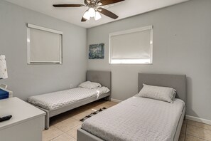 Quarto com duas camas de solteiro, um ventilador de teto, um quadro e uma janela com persianas.