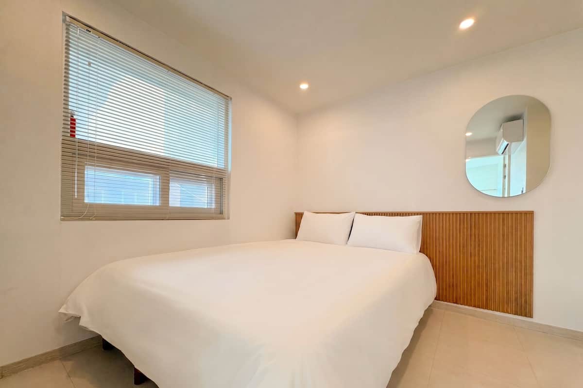 Quarto com cama, janela com persianas, espelho redondo e cabeceira de madeira.