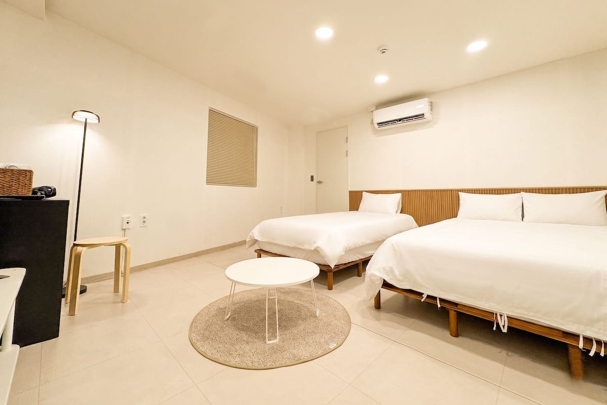 Quarto de hotel com duas camas, uma mesa redonda pequena, abajur, escrivaninha e janela com persianas.
