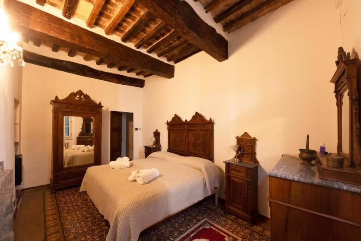 Quarto com cama, cabeceira de madeira, espelho, cômoda e tapete estampado.