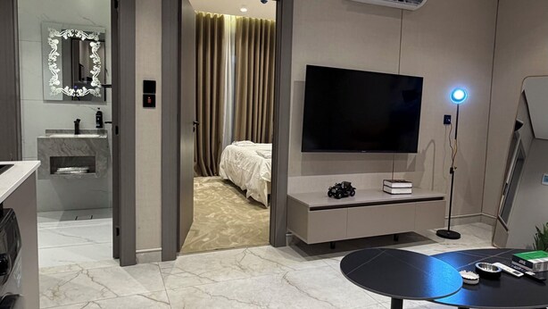 Quarto de hotel moderno com televisão, escrivaninha, cadeira, espelho e banheiro visível através de uma porta aberta.