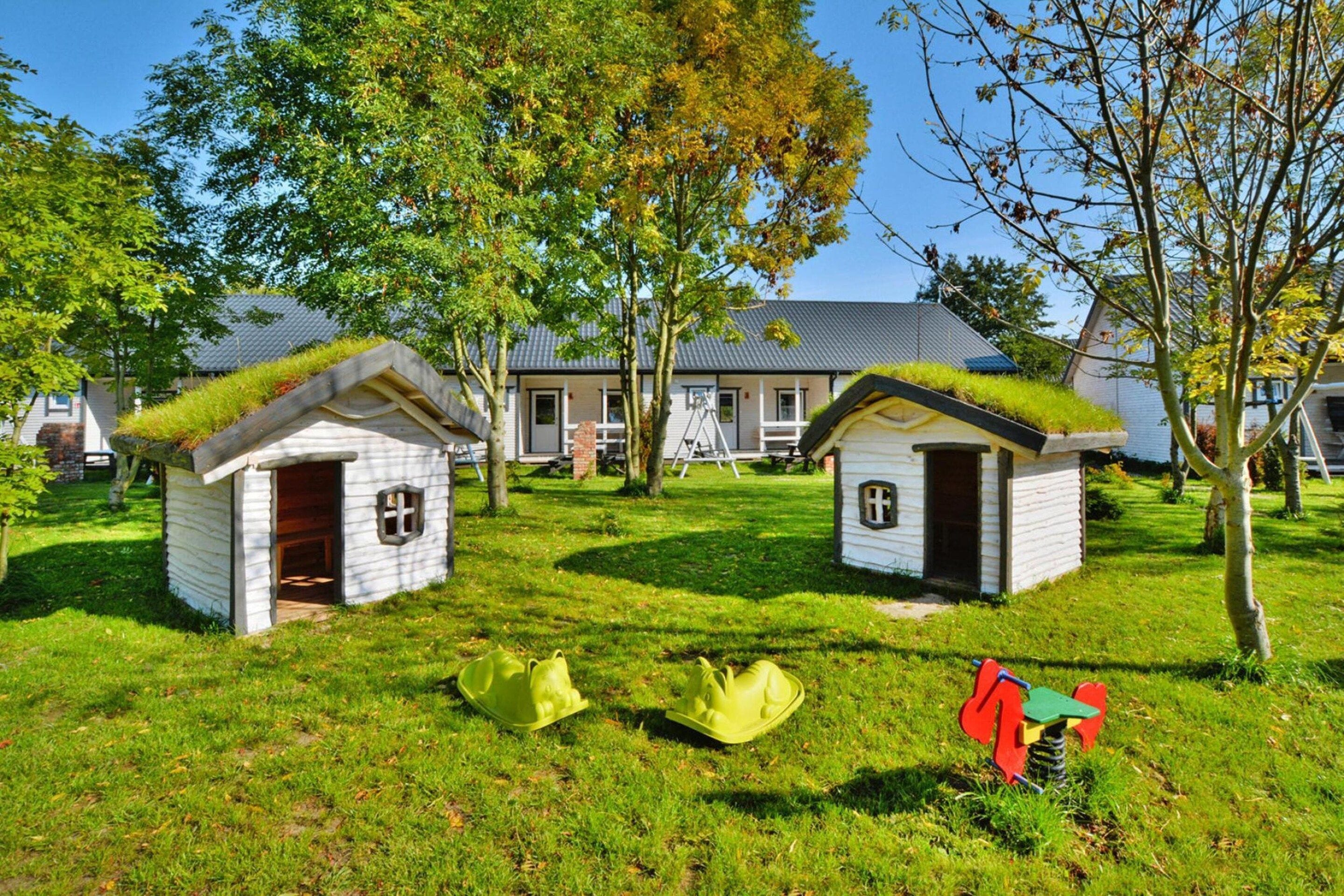 Uma área gramada com dois pequenos edifícios brancos e telhados verdes, uma árvore e um objeto vermelho no gramado.