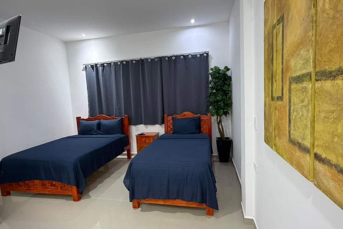 Quarto com duas camas, uma mesinha de cabeceira, um vaso com planta e um quadro abstrato grande na parede.