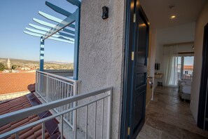 Uma varanda com vista para uma cidade distante, uma porta que dá para um quarto com uma cama e uma televisão, e um chão de azulejos.