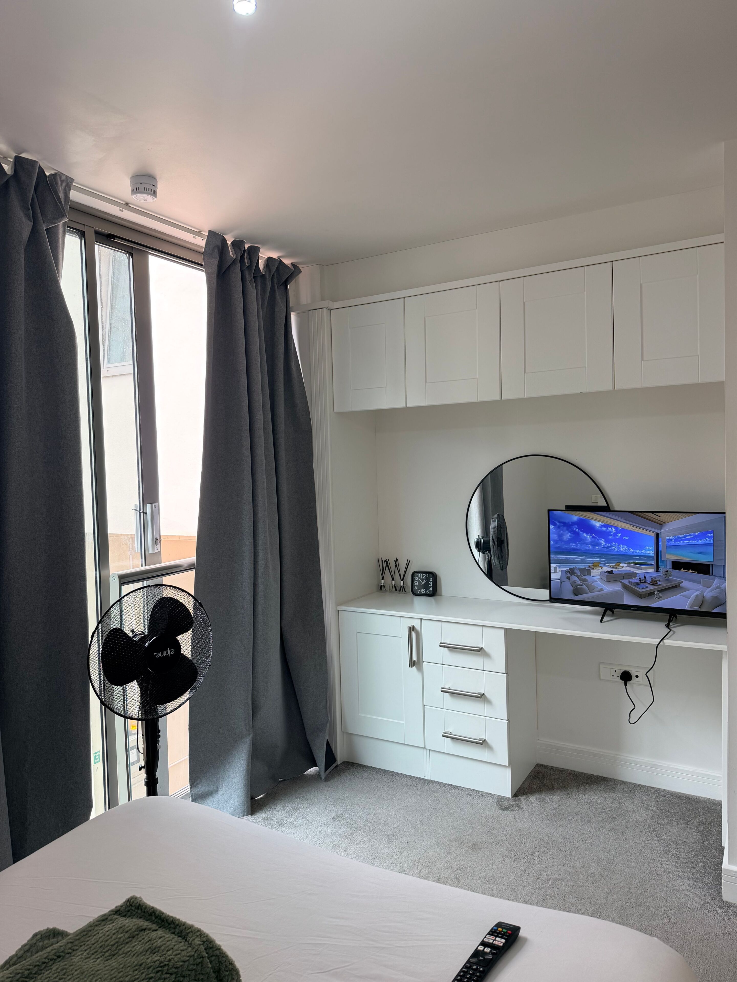Quarto de hotel com uma cama, uma escrivaninha com televisão, um ventilador e uma janela com cortinas.
