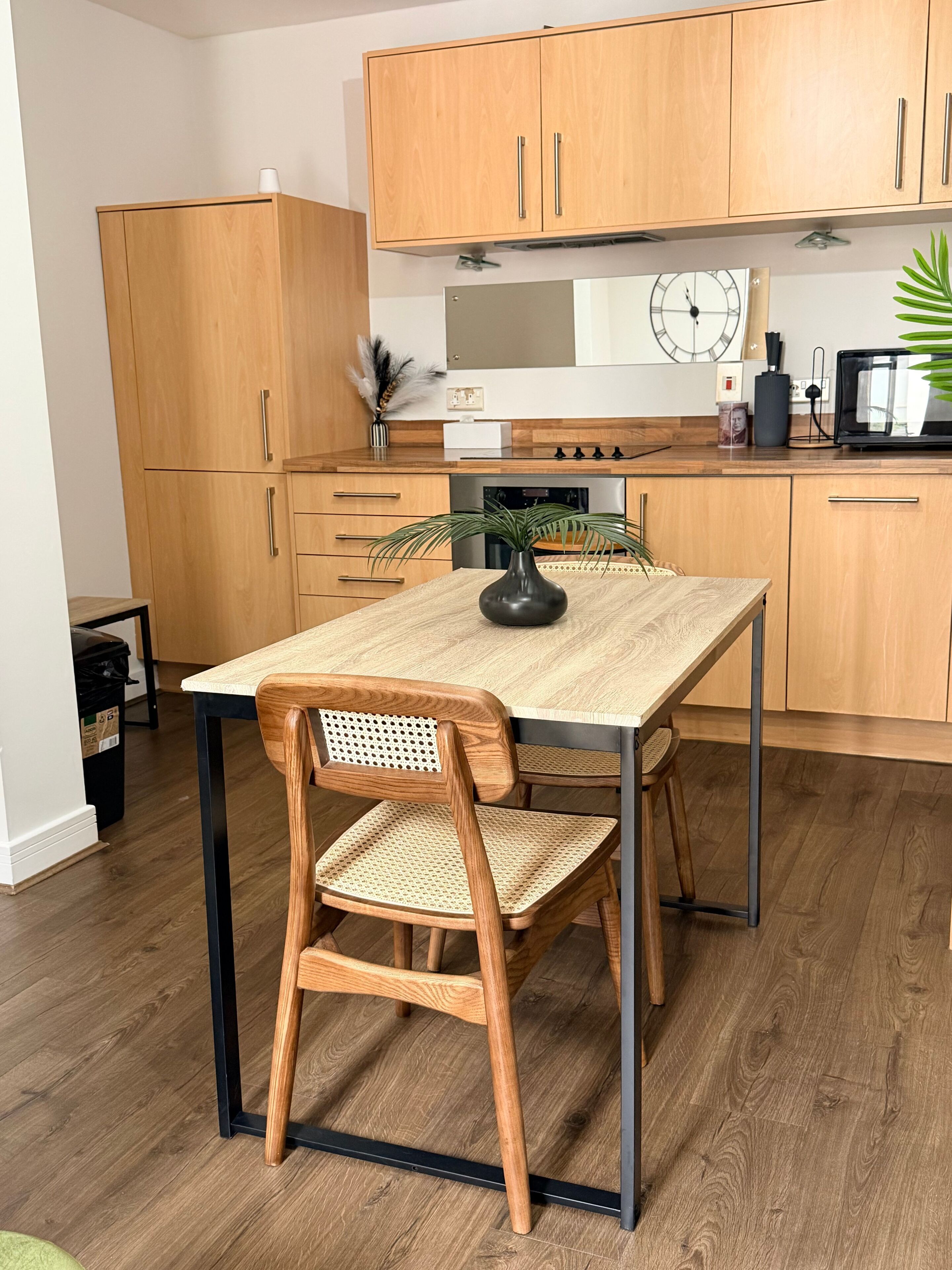 Uma cozinha com armários de madeira, micro-ondas, relógio e um vaso de planta.
