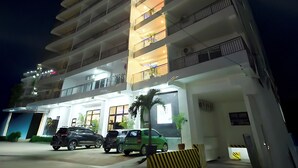 Um hotel com várias andares, varandas, área de estacionamento e um carro verde estacionado na frente.