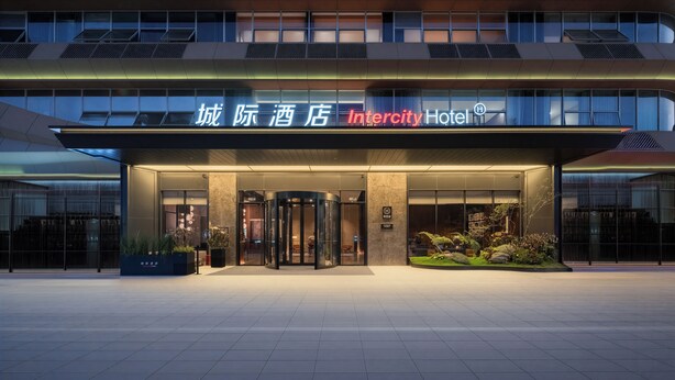 Entrada moderna de hotel com fachada de vidro, porta giratória e placa com a inscrição 'Intercity Hotel'.