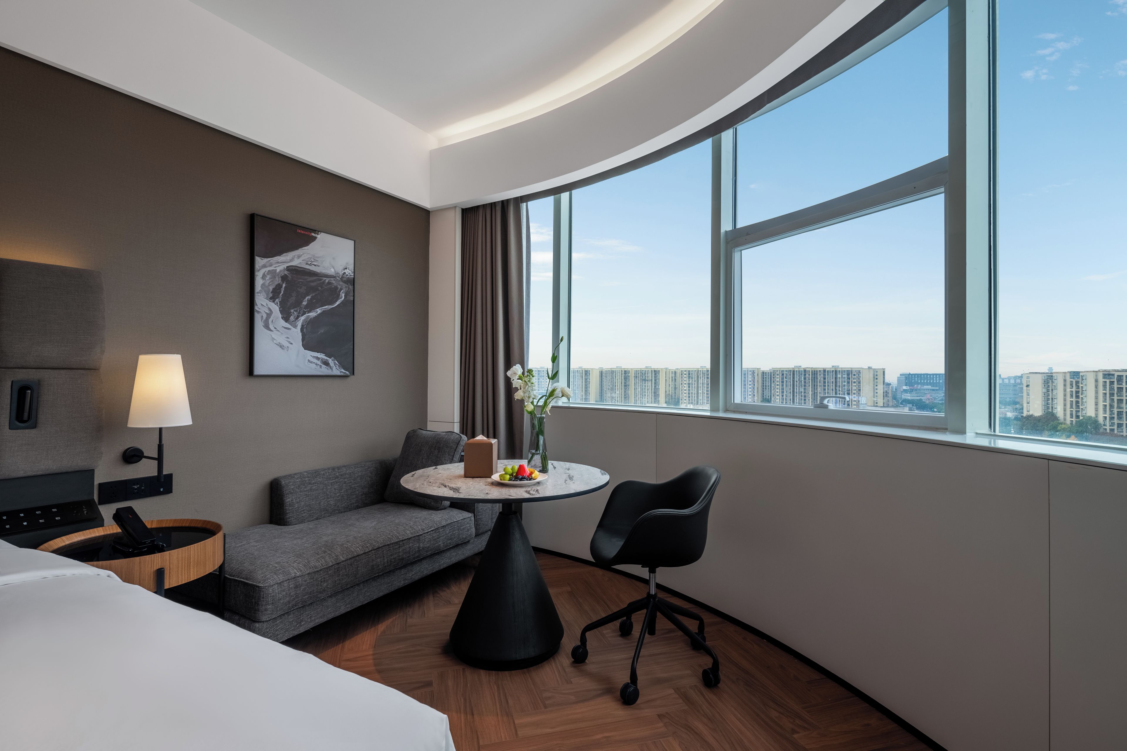Quarto de hotel moderno com uma janela ampla, um sofá, uma mesa redonda pequena com frutas e uma escrivaninha com abajur.