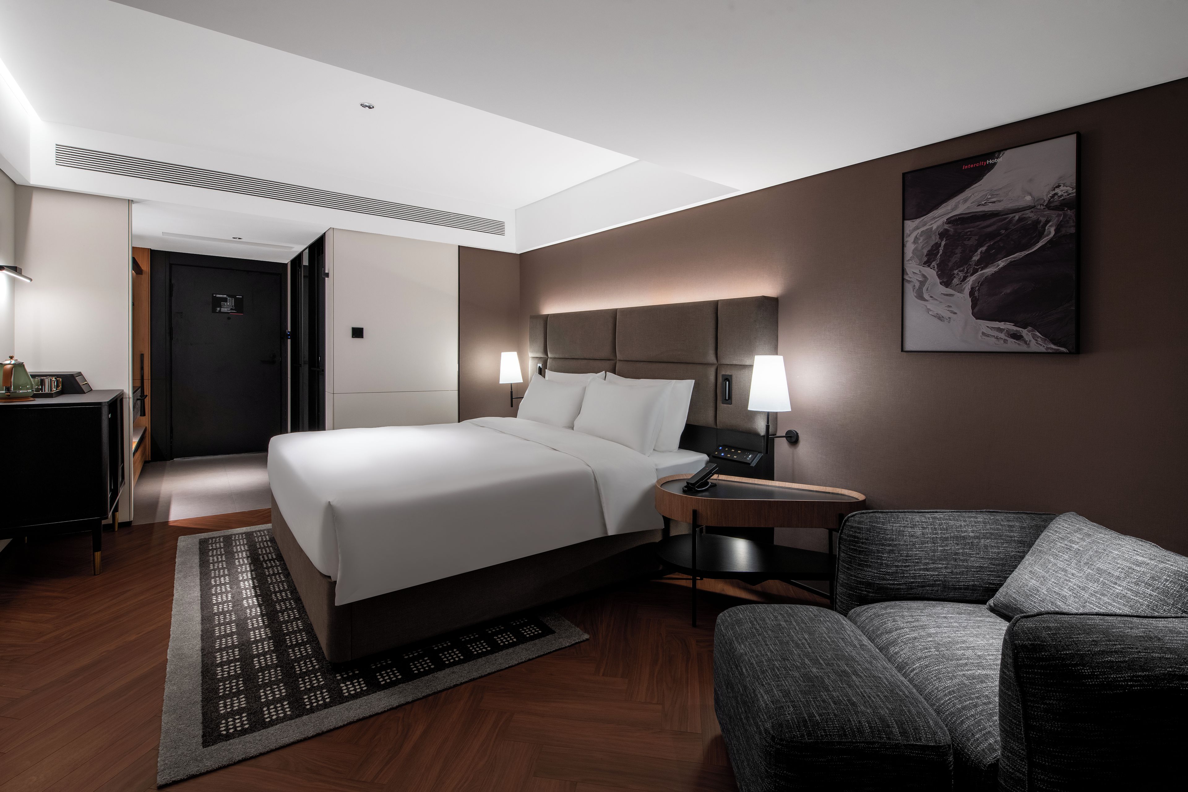 Quarto de hotel moderno com cama, mesa de cabeceira, luminária e uma cadeira.