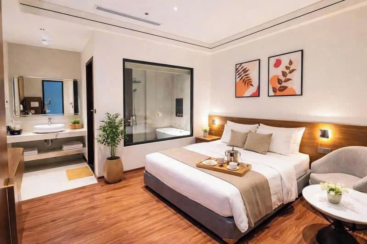 Quarto de hotel moderno com uma cama grande, mesa de cabeceira, cadeira, mesa redonda e banheiro com espelho e pia.