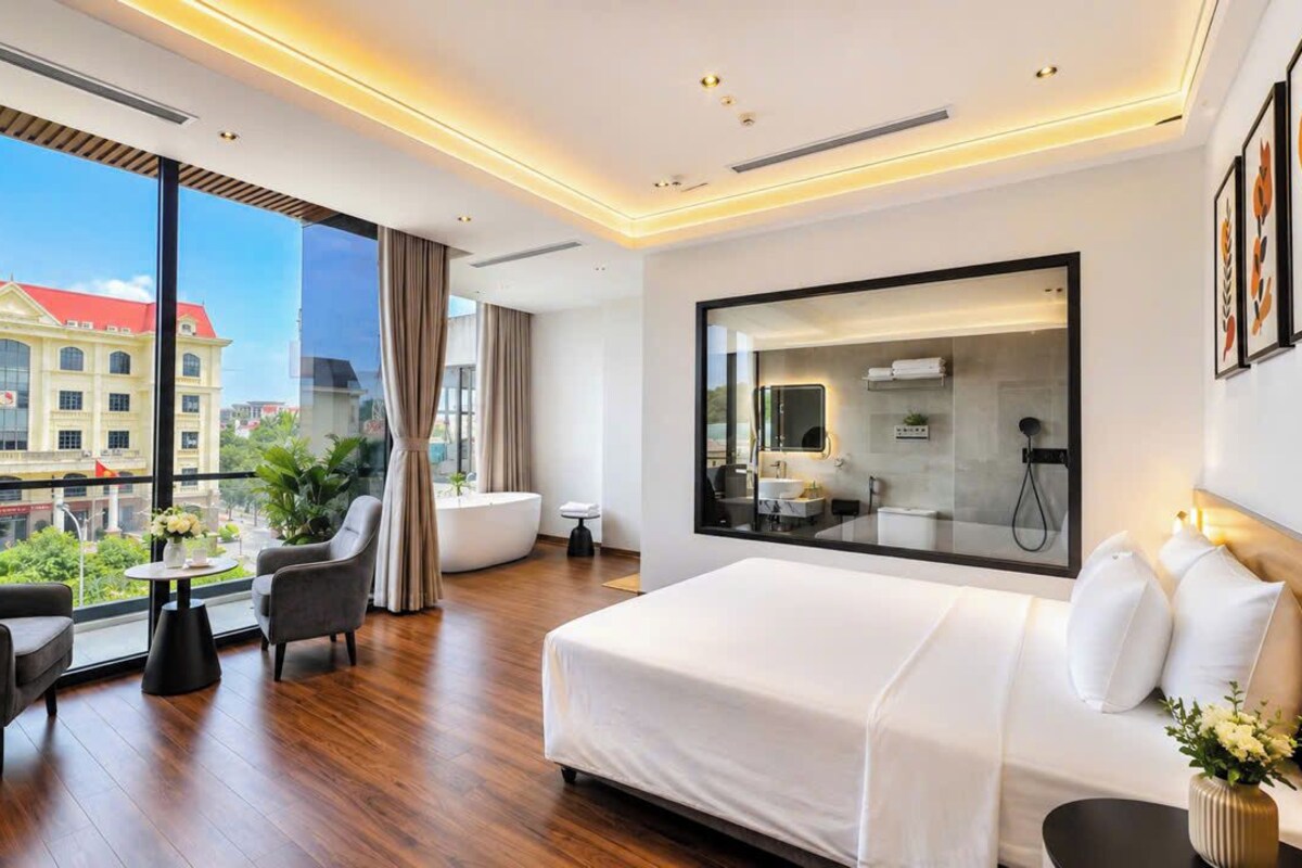 Quarto de hotel com uma cama grande, um espaço de estar com mesa e vista para um edifício pela janela.