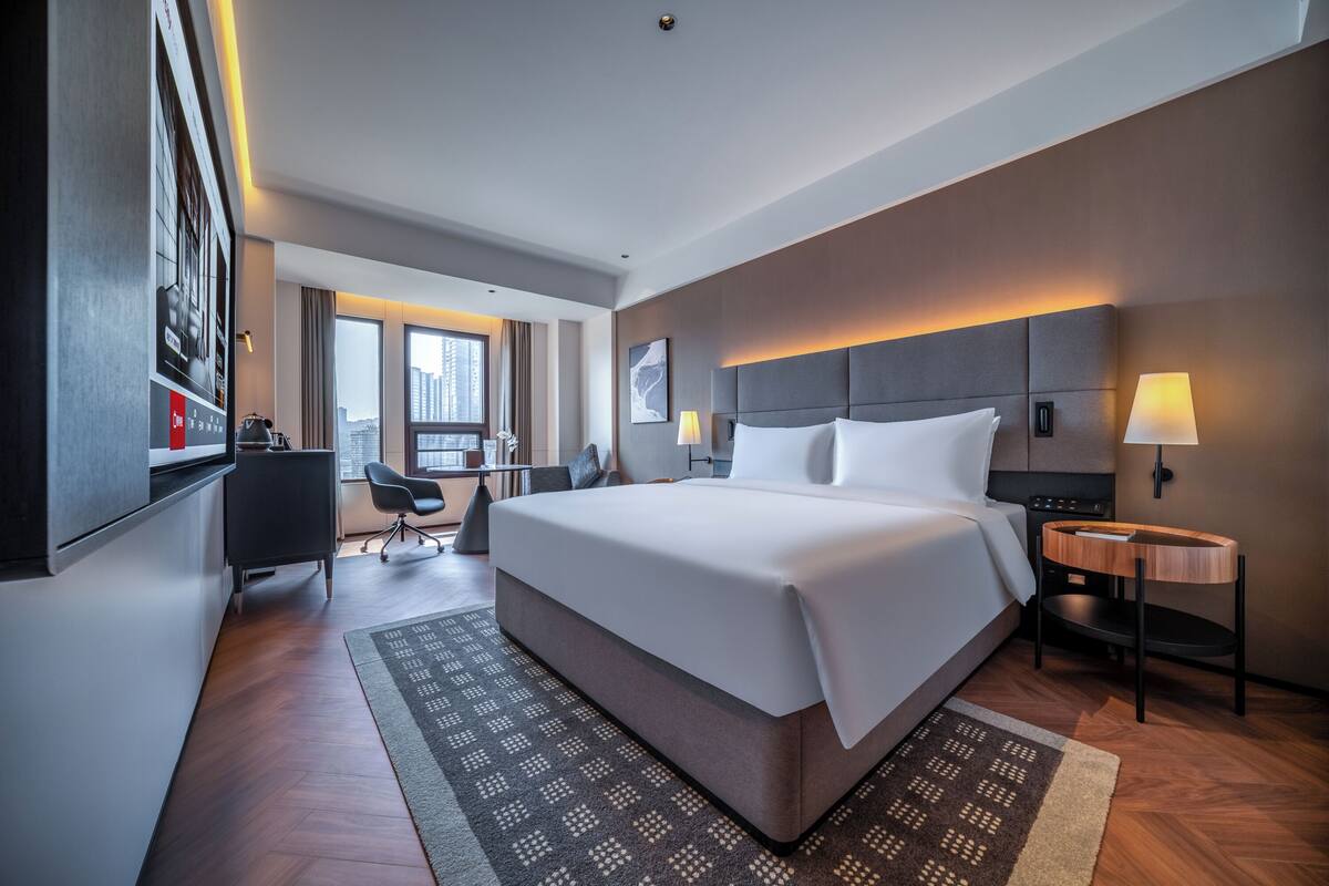 Quarto de hotel moderno com uma cama grande, uma mesa, uma cadeira e vista para a cidade.