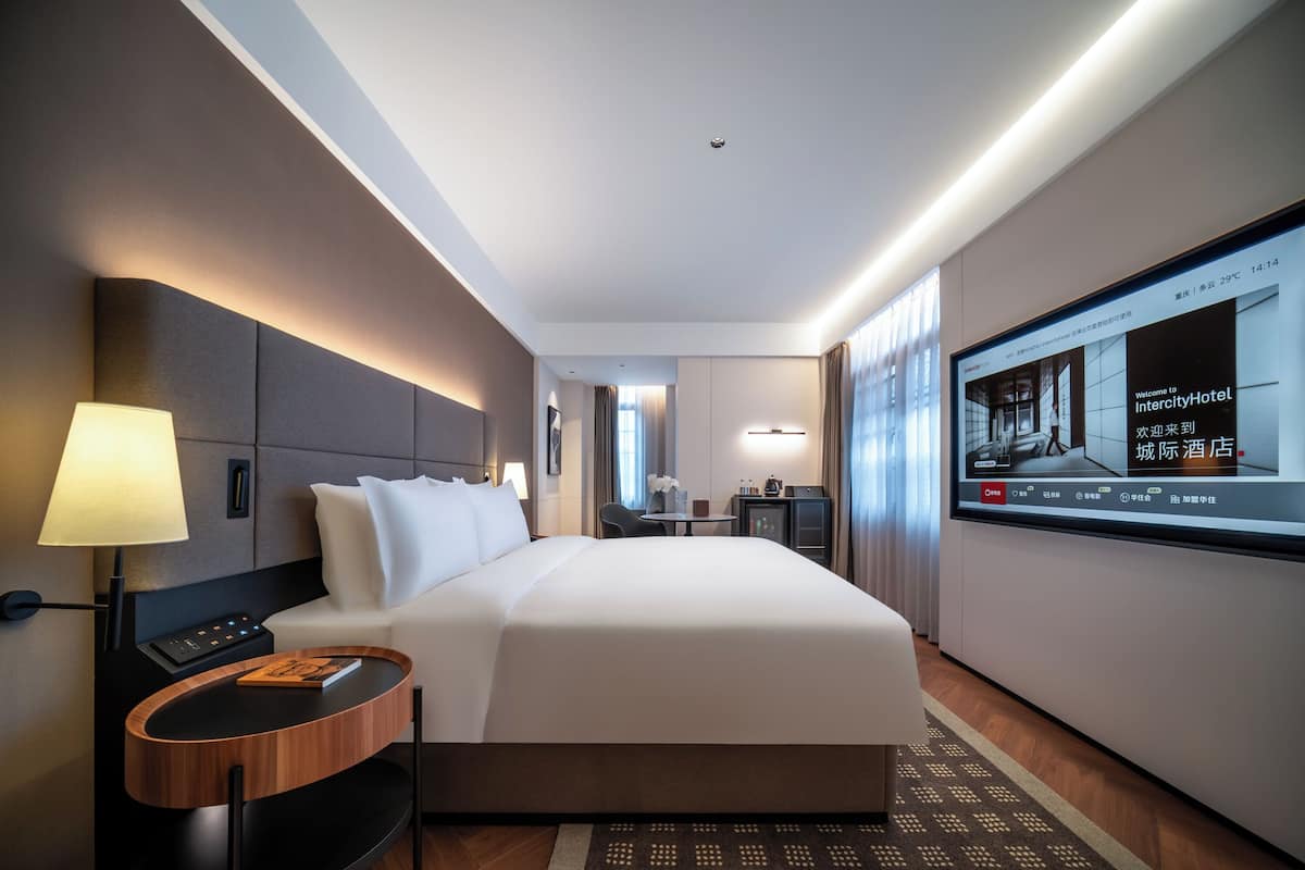 Quarto de hotel moderno com uma cama grande, mesa de cabeceira, luminária e uma televisão exibindo uma mensagem de boas-vindas.