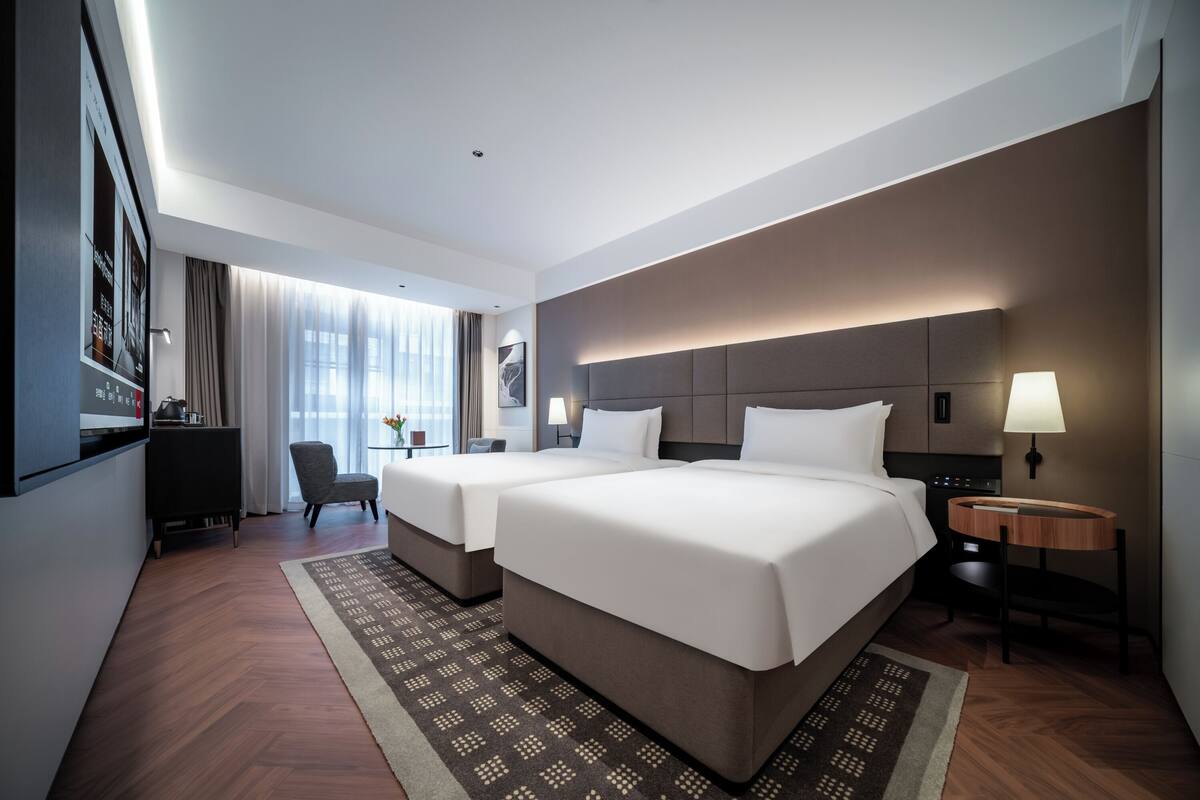 Quarto de hotel com duas camas, uma escrivaninha, uma cadeira, televisão e janela com cortinas.