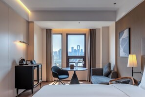 Quarto de hotel com cama, escrivaninha com luminária, cadeira, mesinha com um vaso e vista para a cidade através de grandes janelas.