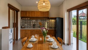 Uma área de refeições com mesa de madeira posta para quatro pessoas, cozinha moderna ao fundo e uma janela grande com vista para o verde.