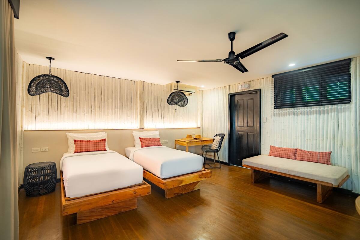Quarto com duas camas, um banco, uma mesa, uma cadeira e um ventilador de teto.