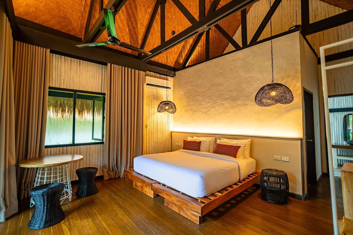 Quarto com estrutura de cama de madeira, uma mesa redonda pequena e dois banquinhos de vime.