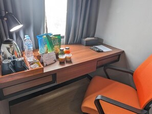 Mesa de quarto de hotel com luminária, garrafa de água, lanches e uma cadeira.
