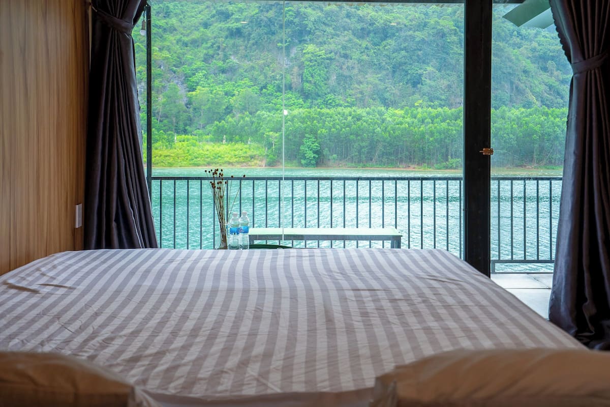 Uma cama com roupa de cama listrada, um banco do lado de fora e vista para uma área arborizada através de um corrimão.
