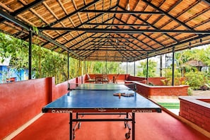Mesa de pingue-pongue em ambiente interno com piso vermelho, cercada por assentos e uma área coberta com telhado de madeira.