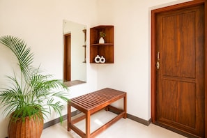 Um banco de madeira, um vaso de planta, uma porta de madeira e uma prateleira fixada na parede com objetos decorativos.