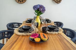 Uma mesa de jantar de madeira com pratos pretos, um vaso amarelo com flores roxas e um prato de pitaya e mamão.