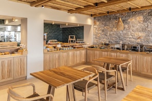 Um café com mesas e cadeiras de madeira, parede de pedra e área de cozinha ao fundo.