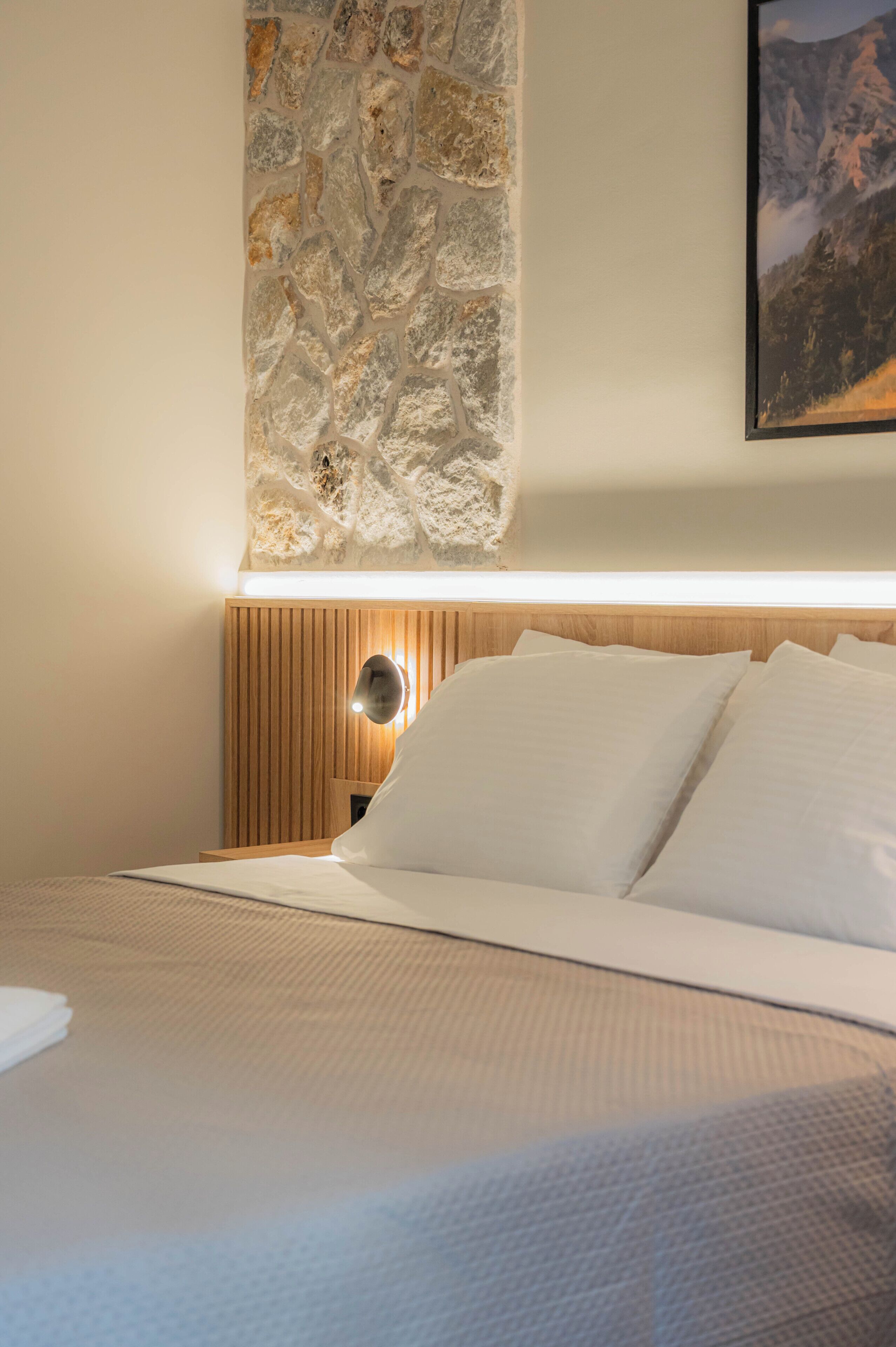 Quarto de hotel com parede de pedra, cabeceira de madeira, cama com roupa de cama branca e um quadro pendurado na parede.