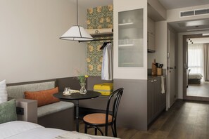 Quarto de hotel compacto com cama, pequena mesa de jantar, sofá, poltrona, cozinha compacta e banheiro visível através de uma porta aberta.