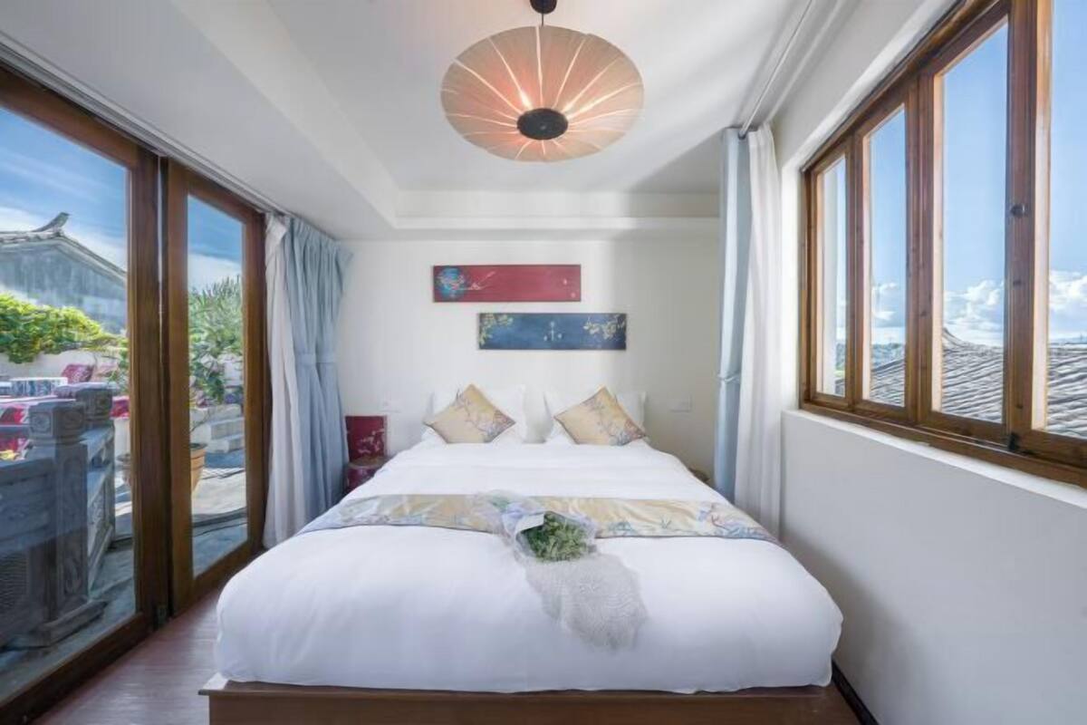 Quarto com cama grande, luminária no teto e vista de um edifício tradicional por grandes janelas.