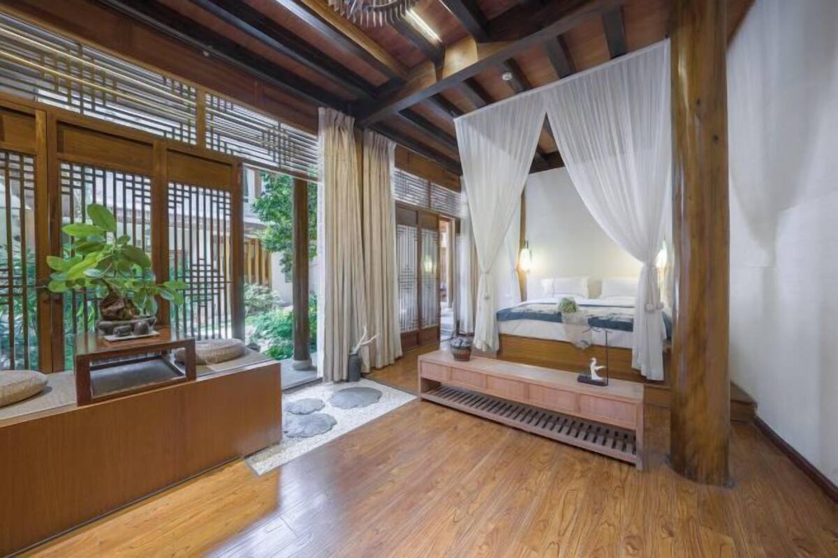 Quarto com piso de madeira, uma cama com cortinas brancas, mesa de cabeceira e um vaso de planta.