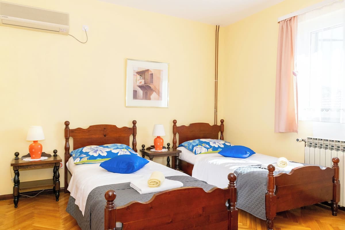 Quarto de hotel com duas camas de madeira, um radiador, uma janela com cortinas e um ar condicionado fixado na parede.